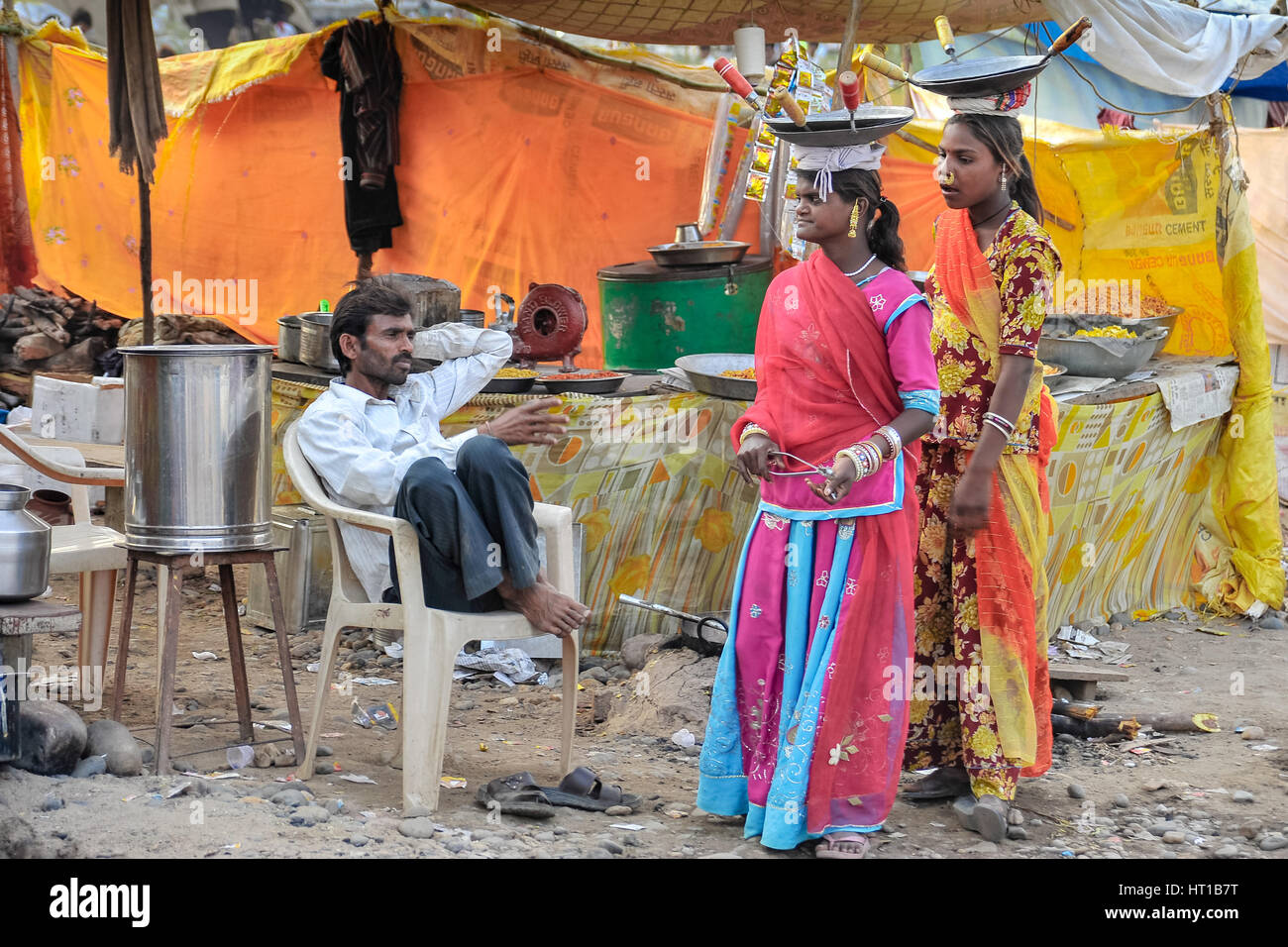 Two young women carry skillets on their heads in front of a chai stall ...
