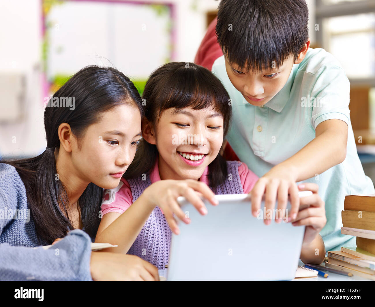 three happy asian elementary school students looking at tablet computer smiling in classroom ...