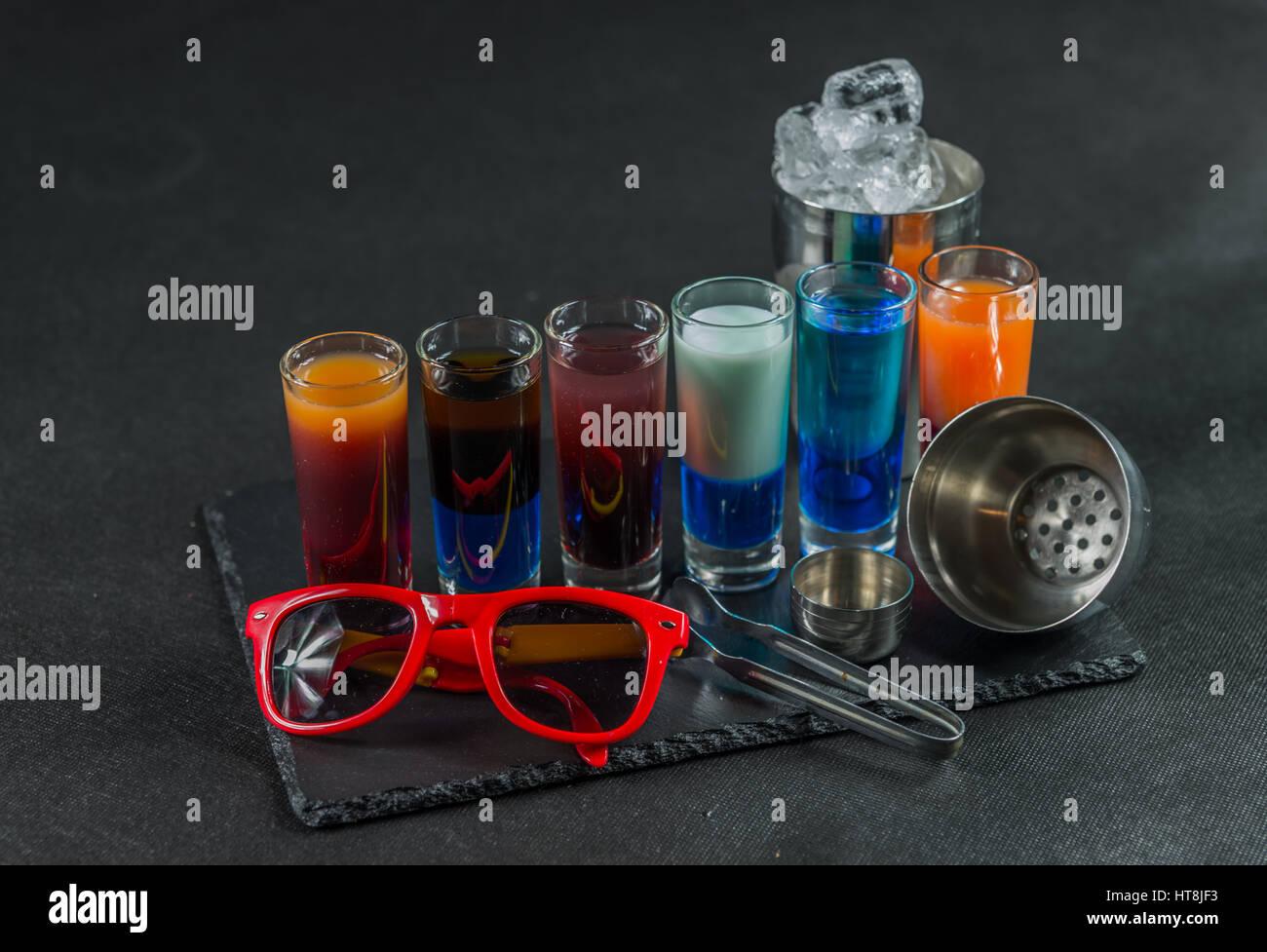six different colored shot drinks, lined up on a black stone plate, ice ...