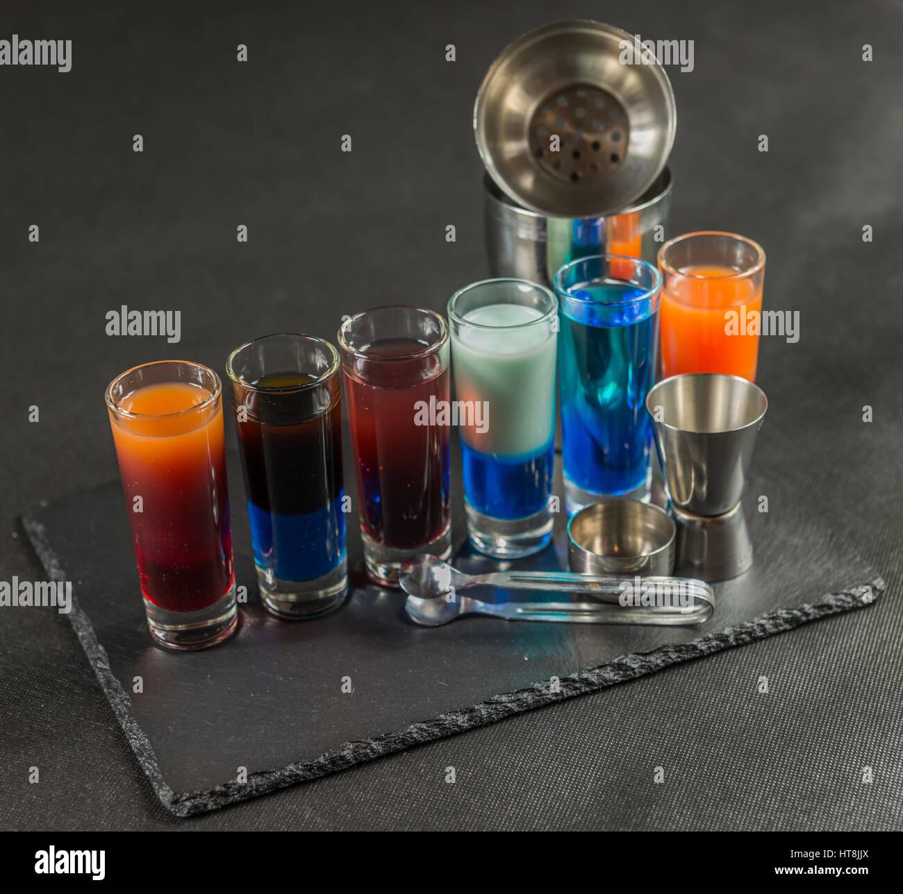 six different colored shot drinks, lined up on a black stone plate, ice ...
