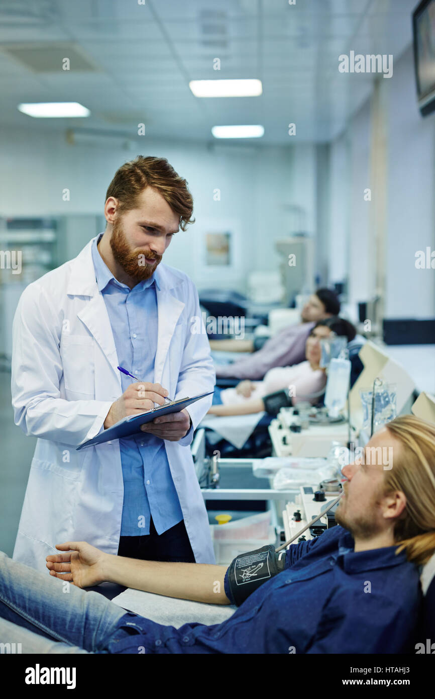 Doctor making notes while talking to young man during hemotransfusion ...
