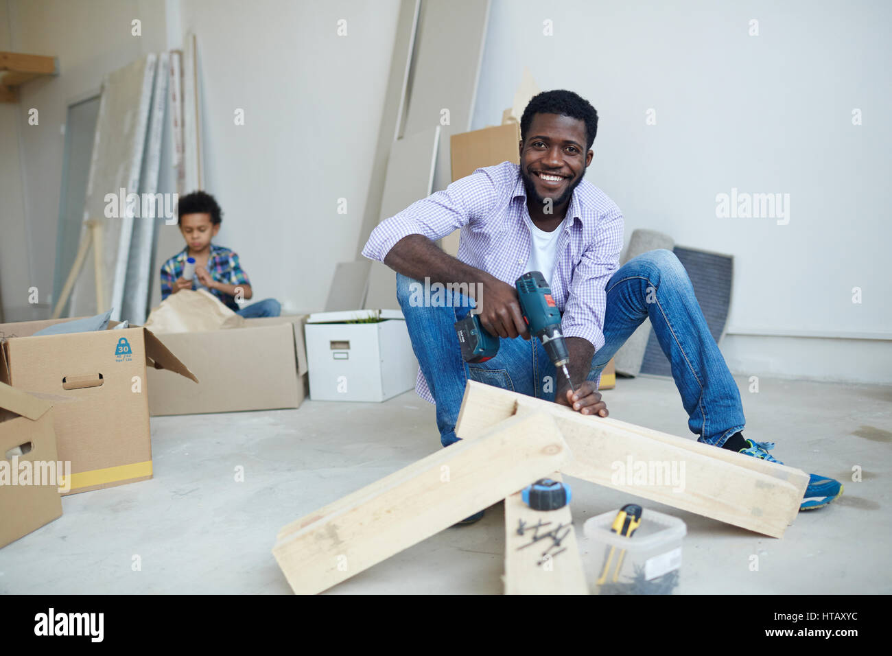 Young man drilling wooden planks while his son unpacking boxes on ...