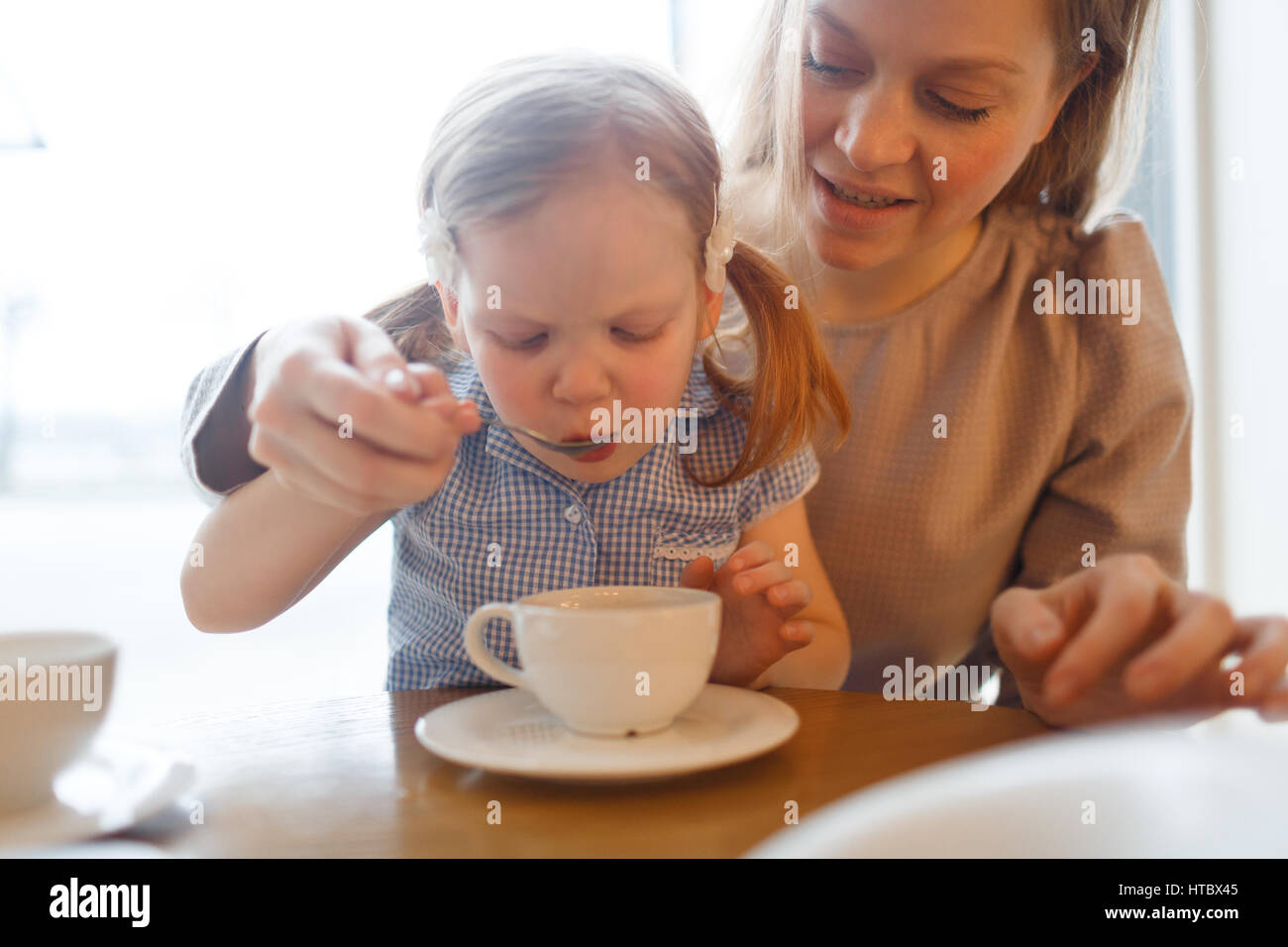Adorable child sipping hot drink from spoon with her mother near by ...
