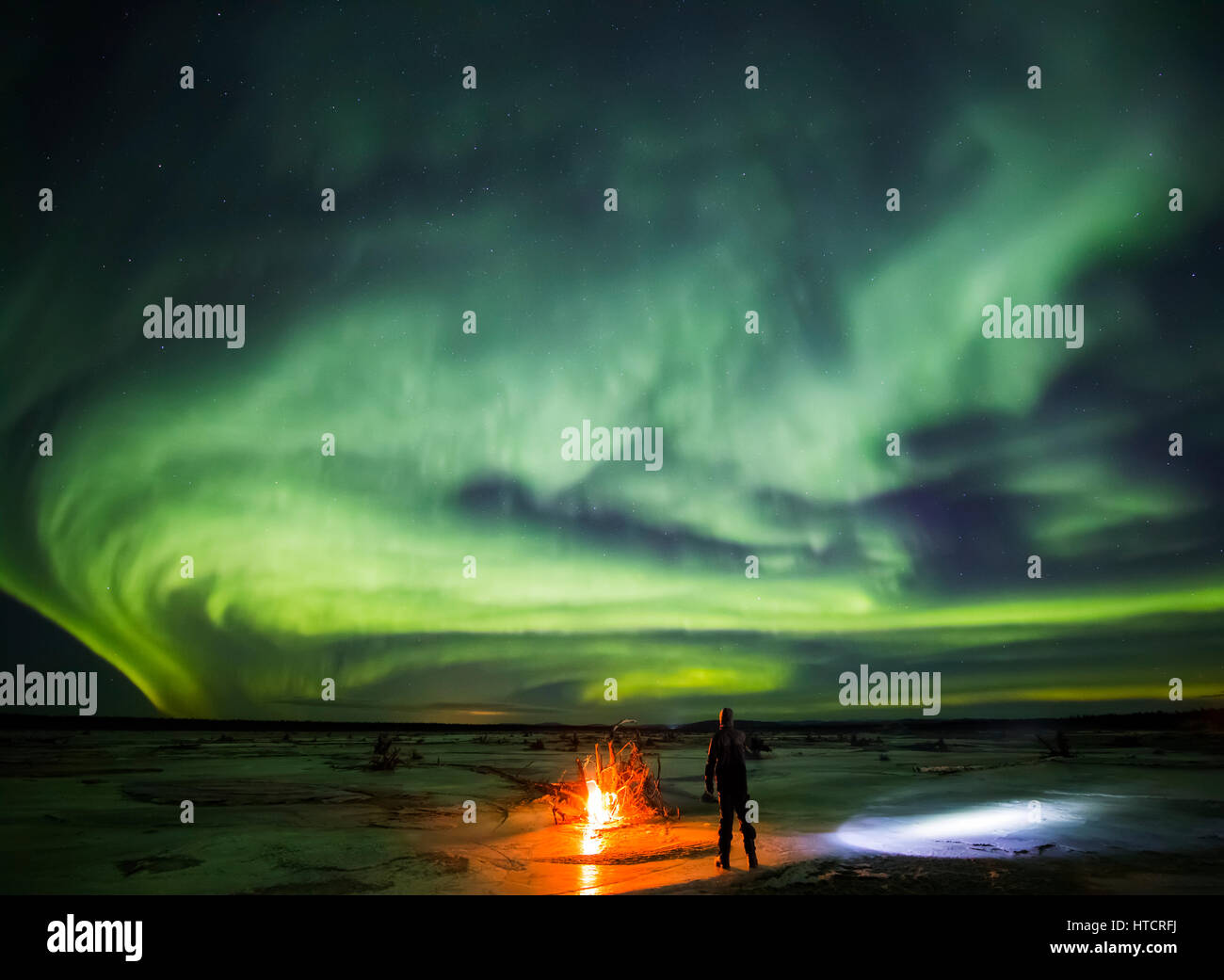 Man stands next to a campfire while northern lights dance overhead ...