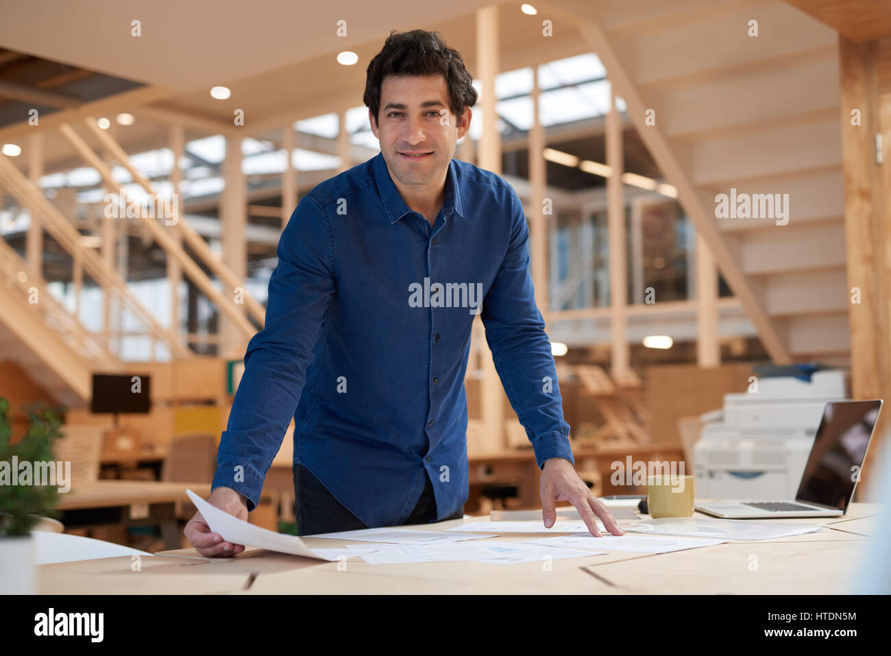 Portrait of a smiling young businessman leaning over a desk in a modern ...
