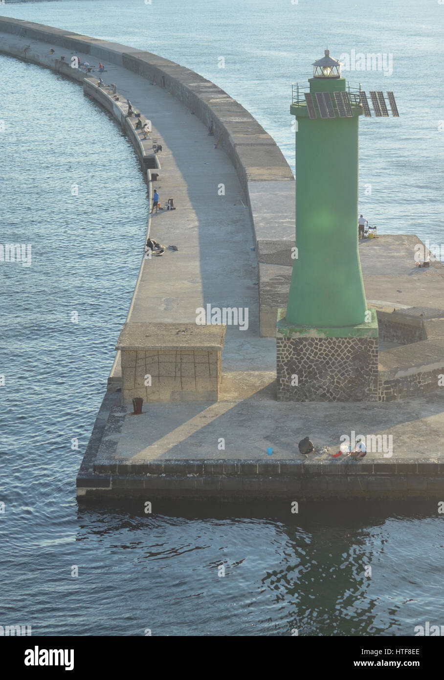 A green light house at the end of a stone pier. Some people are fishing ...