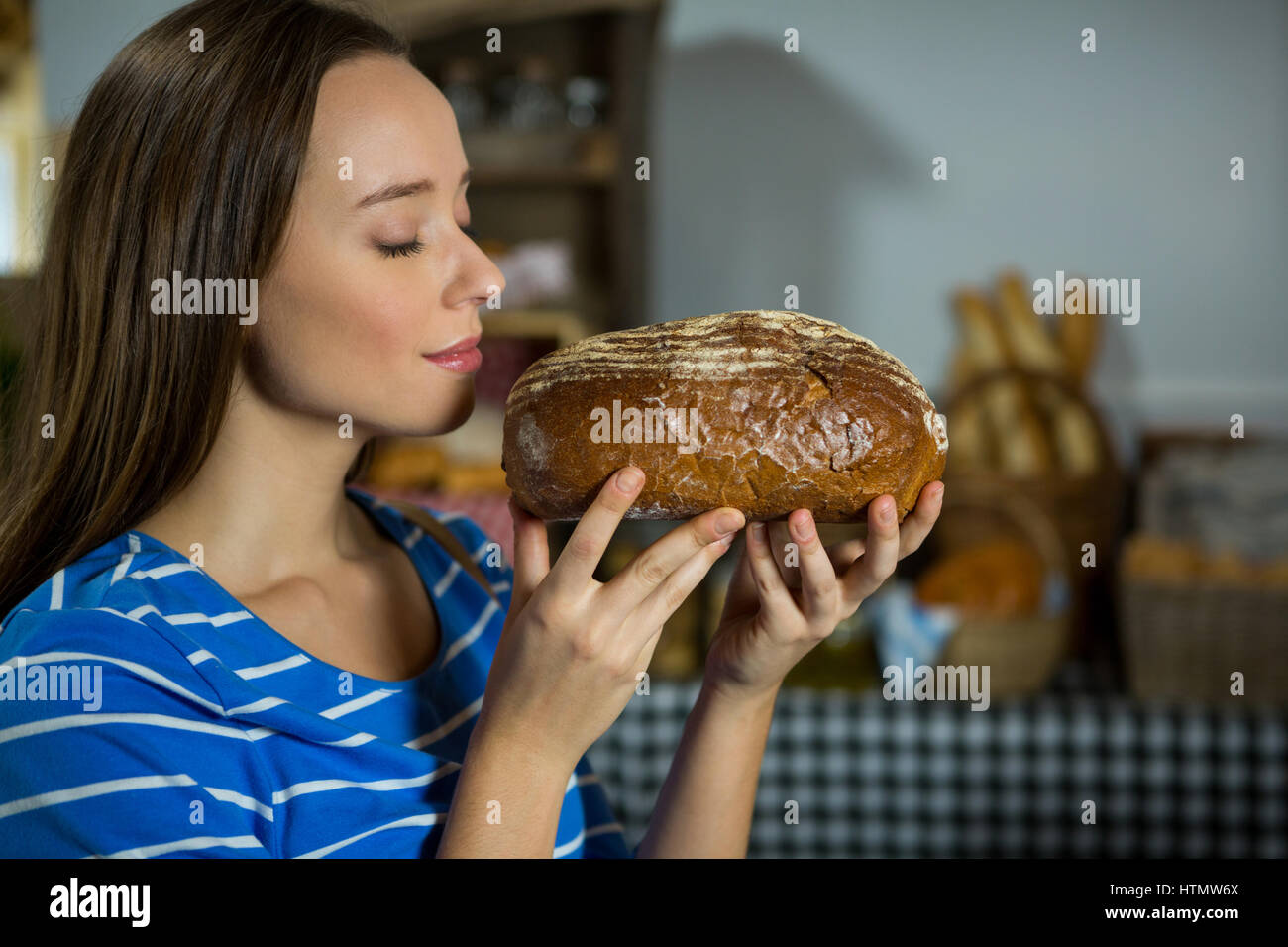 Smiling woman smelling a round loaf of bread at counter in bakery shop ...