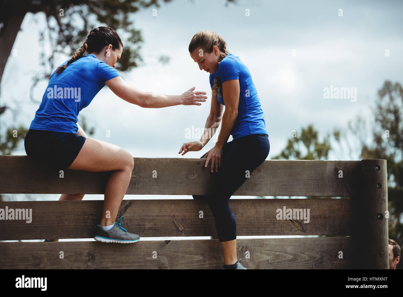 Female trainer assisting woman to climb a wooden wall during obstacle ...