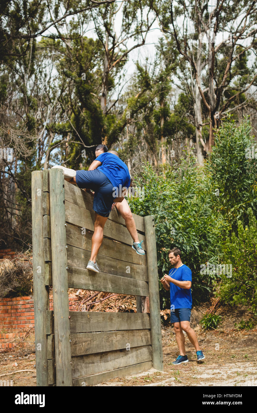 Female trainer assisting man to climb a wooden wall during obstacle ...