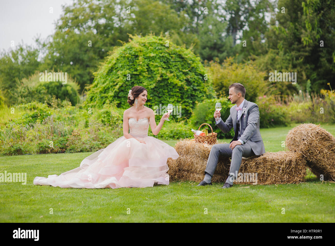 Bride and groom make toast sitting on a bale of hay. Grass, trees and ...