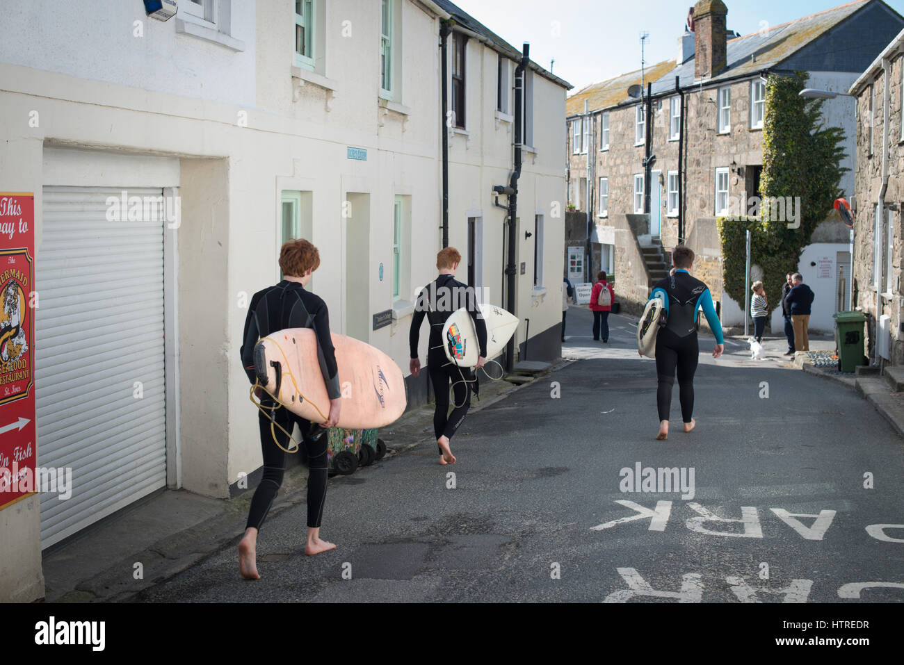 Teenage boys wearing wetsuits carry surf boarders past cottages in St ...