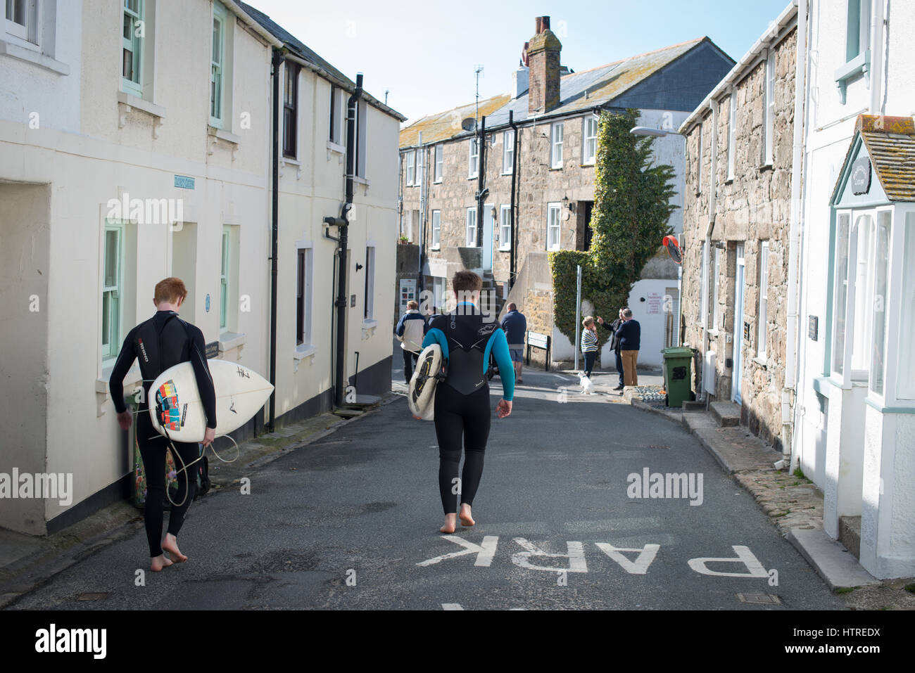 Teenage boys wearing wetsuits carry surf boarders past cottages in St ...