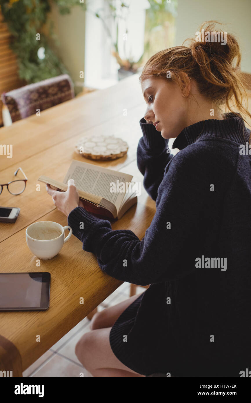 Woman sitting by dining table and reading a book Stock Photo - Alamy