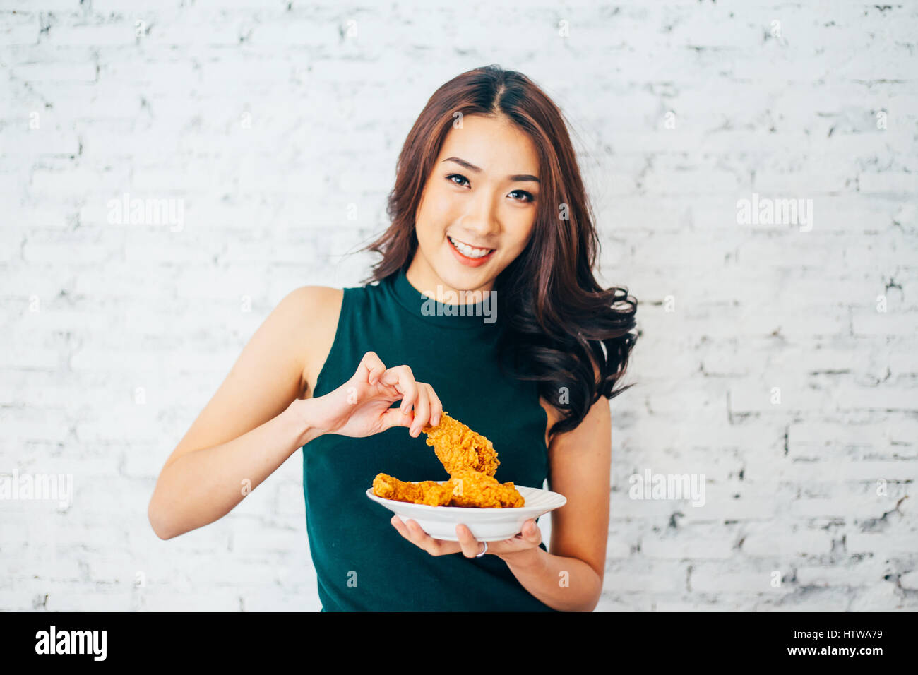 Attractive Asian Woman Eating Fried Chicken Drumstick Over White Brick