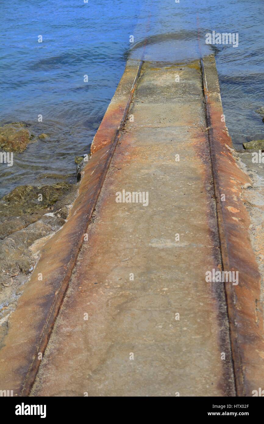 Rusty old iron and stone slipway leading from boathouse to the blue ...