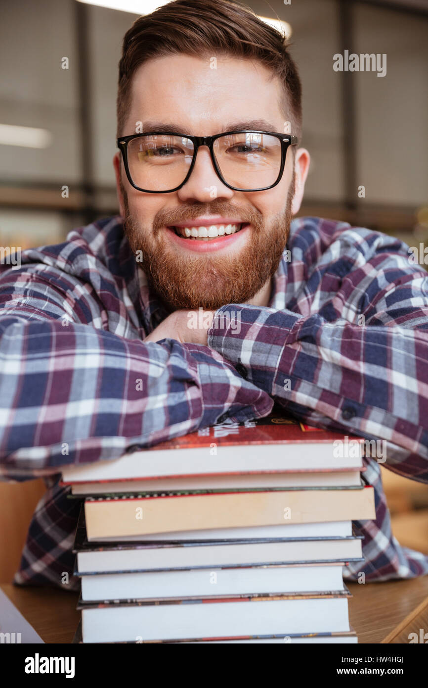 Smiling male student resting on stack of books while sitting at the ...