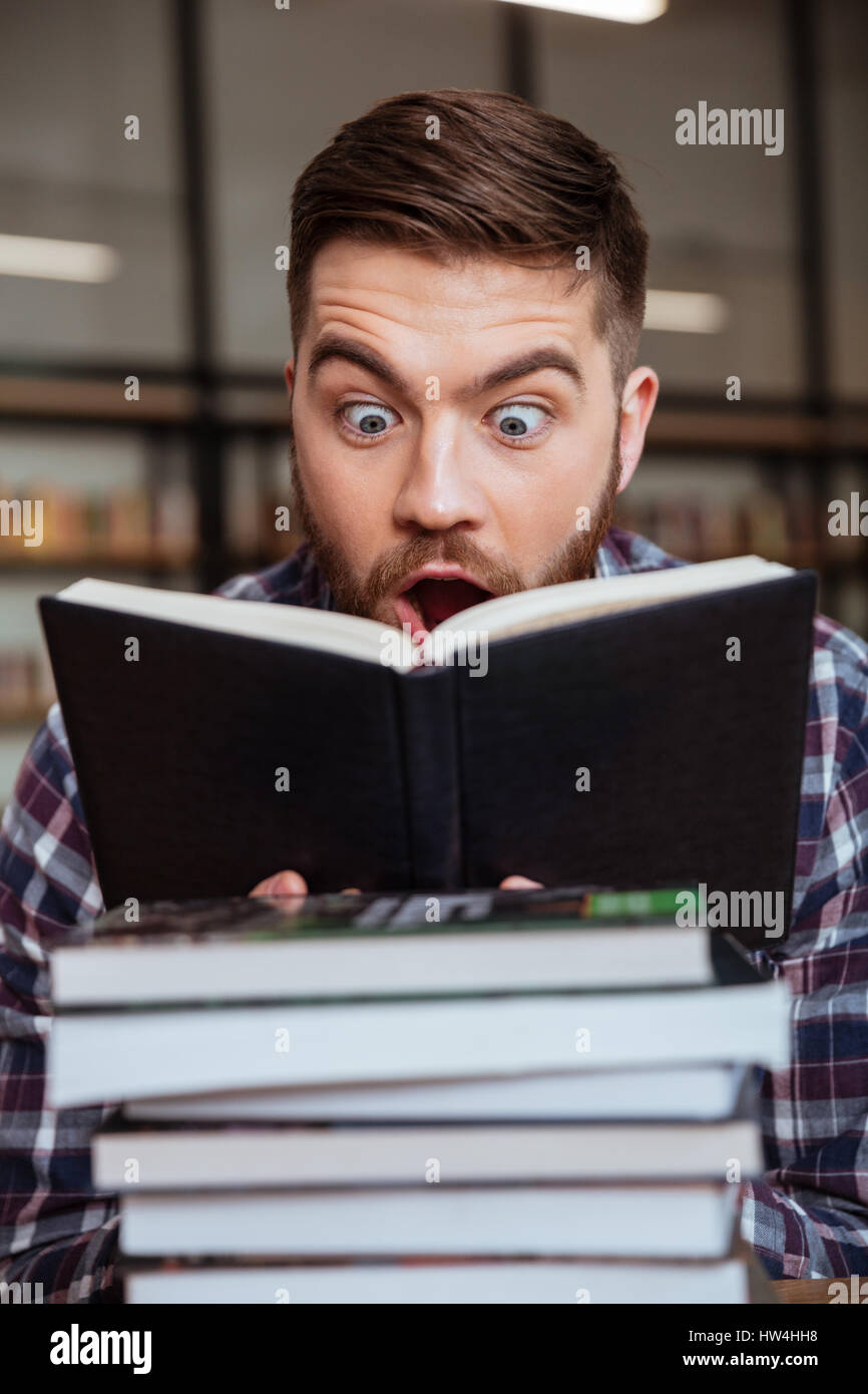 Portrait of surprised male student reading book behind stack of books ...