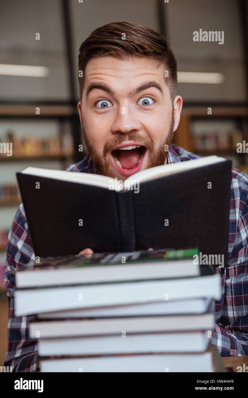 Portrait of excited male student reading book behind stack of books on ...
