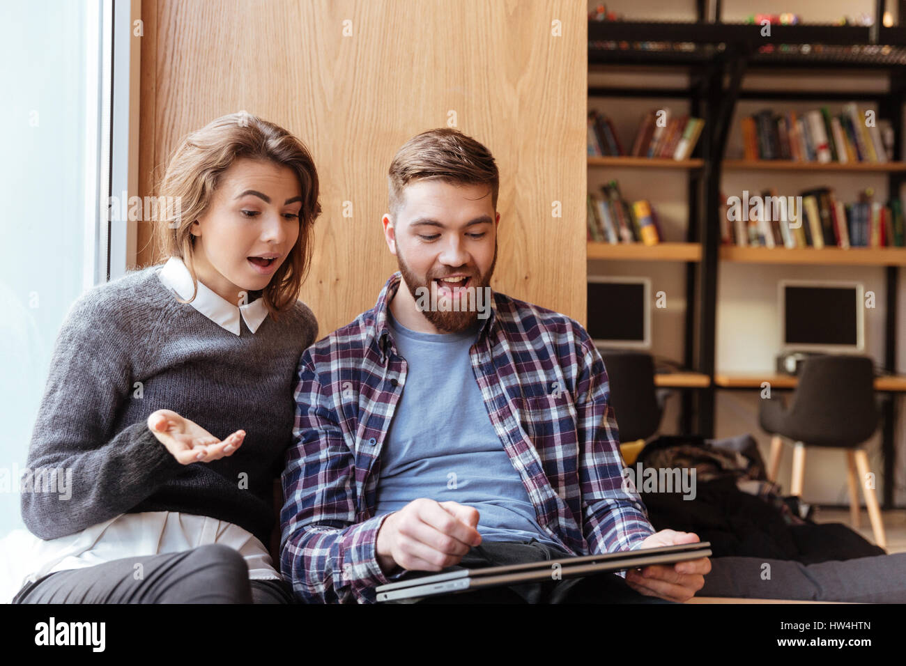Two young students using tablet computer while sitting on a windowsill ...
