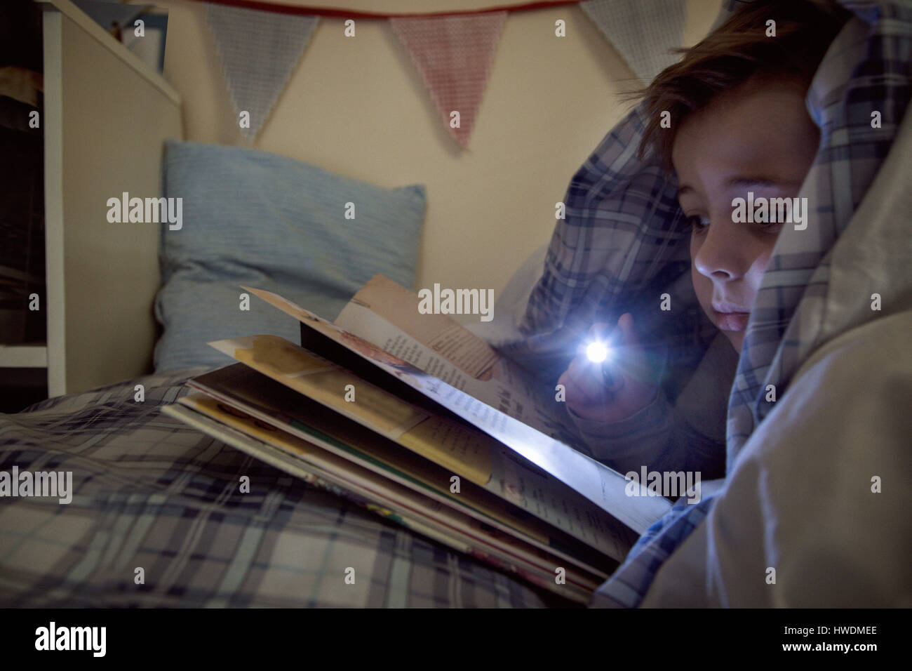 Boy lying in bed wrapped in duvet reading book by torchlight Stock ...