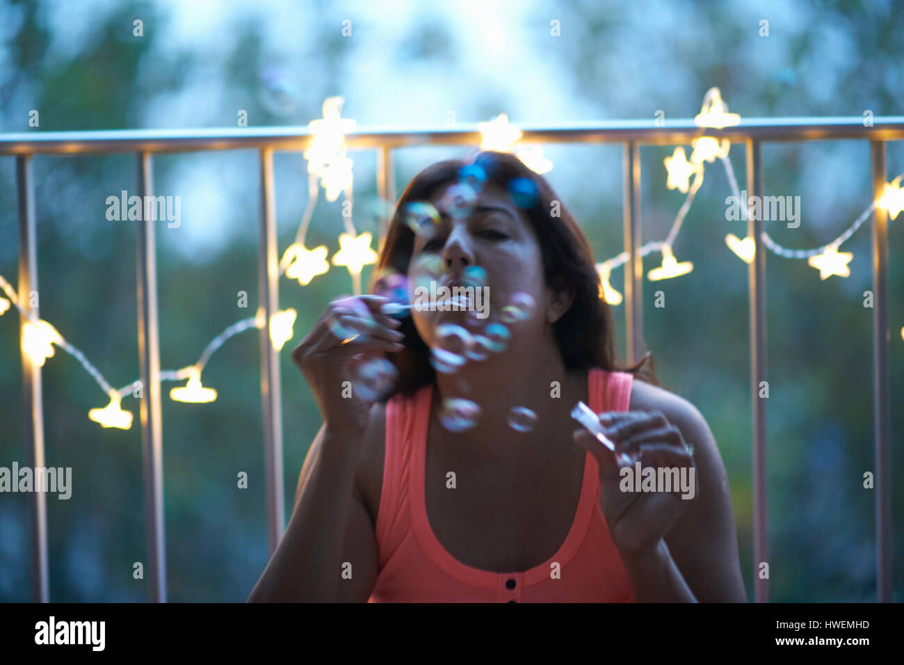 Mature woman blowing bubbles by illuminated decorative lights at dusk ...