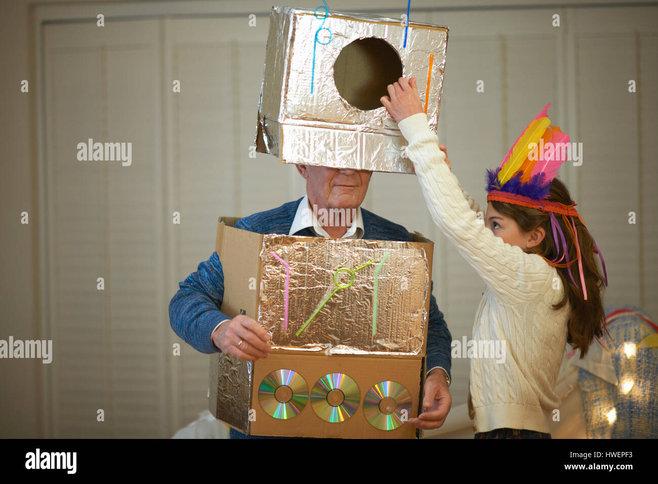 Girl in feather headdress putting robot costume onto grandfather Stock ...