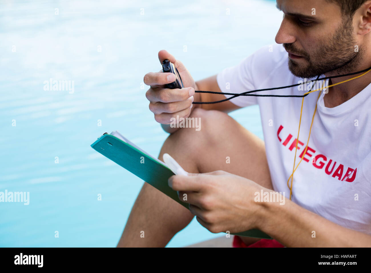 Lifeguard holding clipboard and stopwatch at poolside on a sunny day ...
