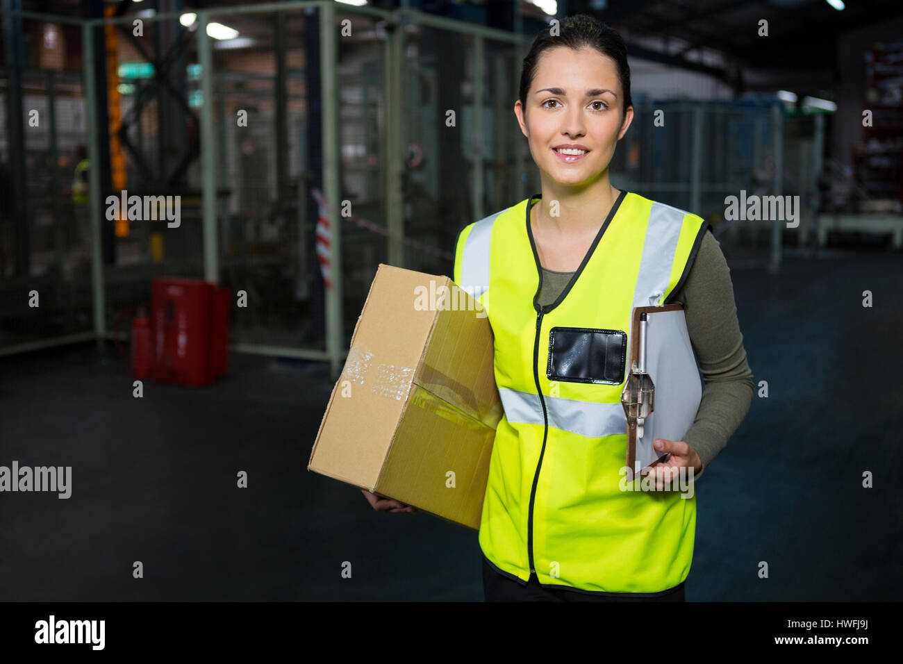 Portrait of young female worker carrying box and clipboard in warehouse ...