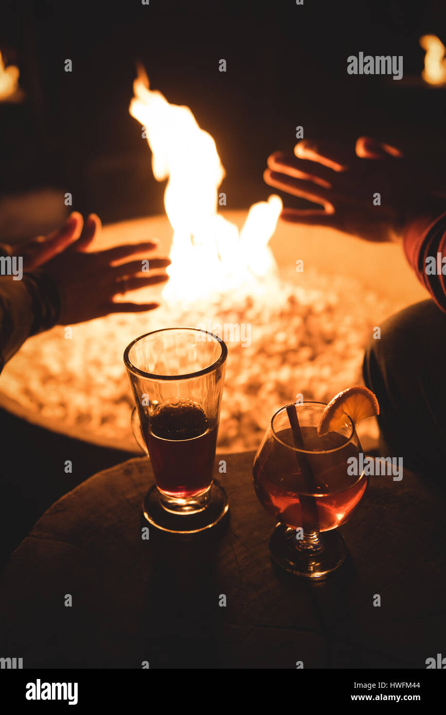 Midsection of couple sitting by fire pit and drinks at night during ...