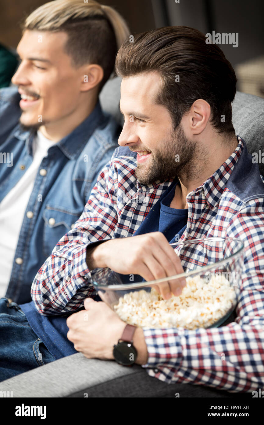 Smiling male friends sitting on couch and eating popcorn Stock Photo ...