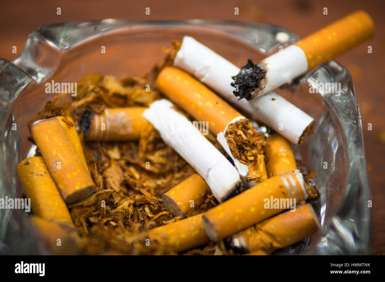 Cigarettes and tobacco lying inside and around glass ash tray on wooden ...