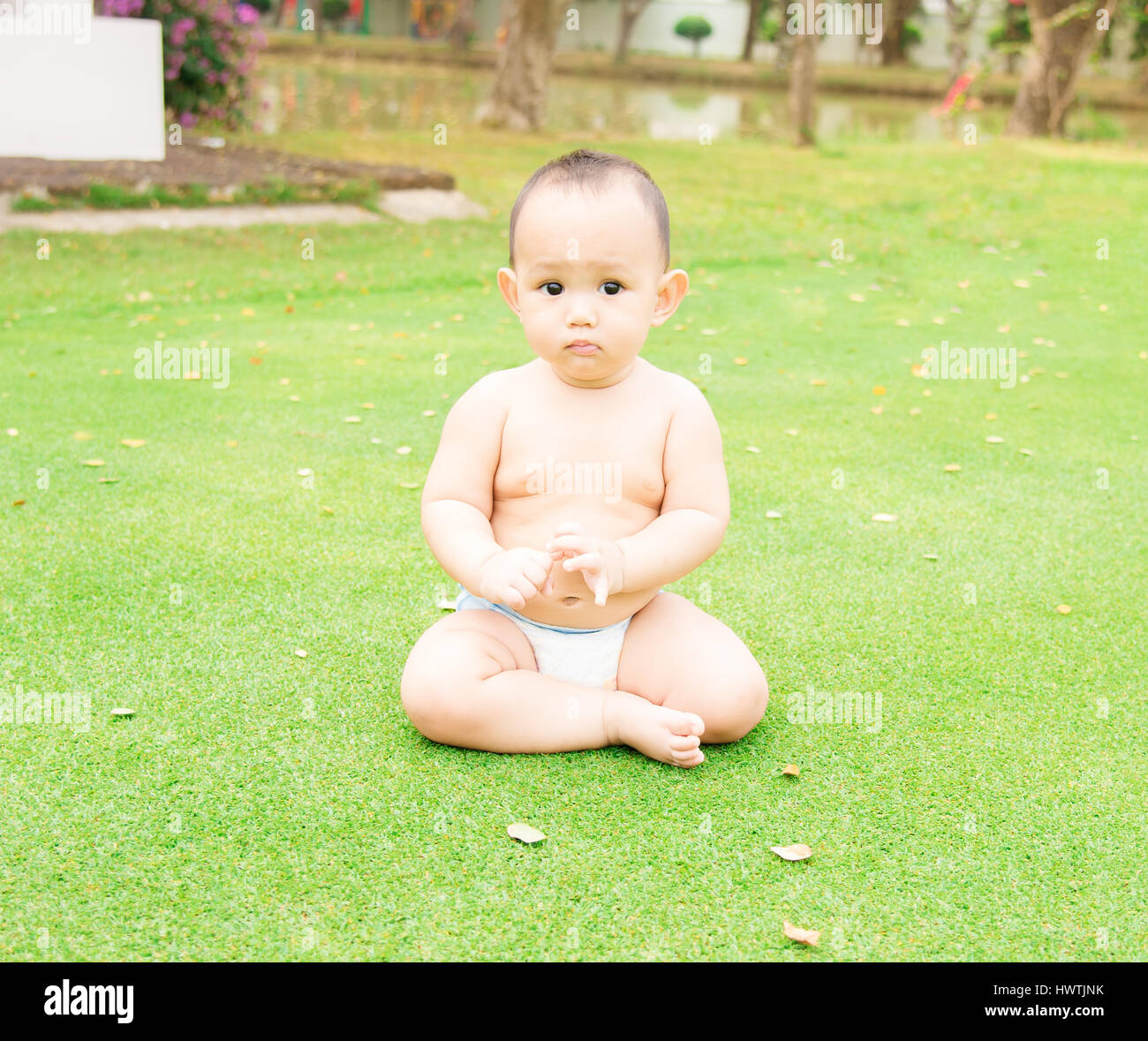 little Thai baby sitting on the green grass field at the outdoor park ...