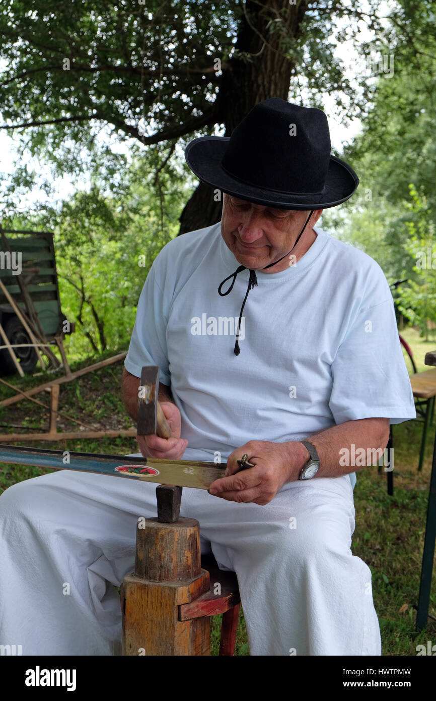 Farmer with hammer and iron tool on the tree stump is sharpening his ...