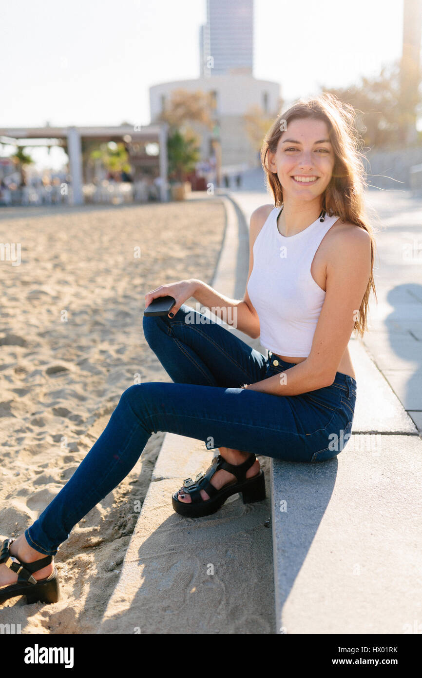 Spain, Barcelona, portrait of smiling young woman sitting on beach ...