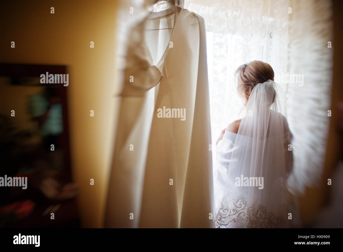 Bride on silk robe looking on window with wedding dress at room on ...
