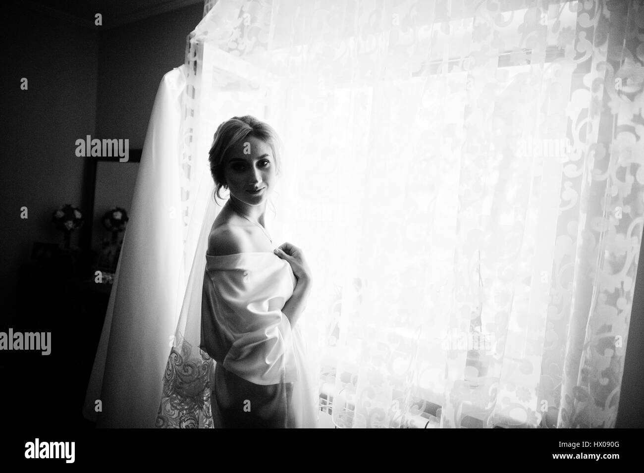 Bride on silk robe looking on window with wedding dress at room on ...