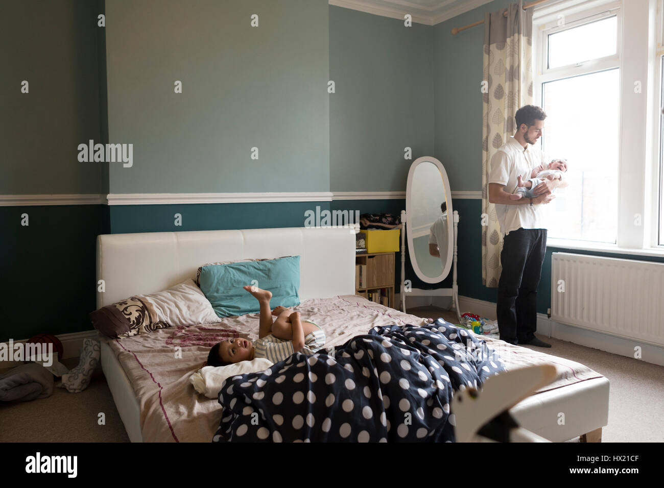 Little boy rolling around on his father's bed, who is standing by the ...