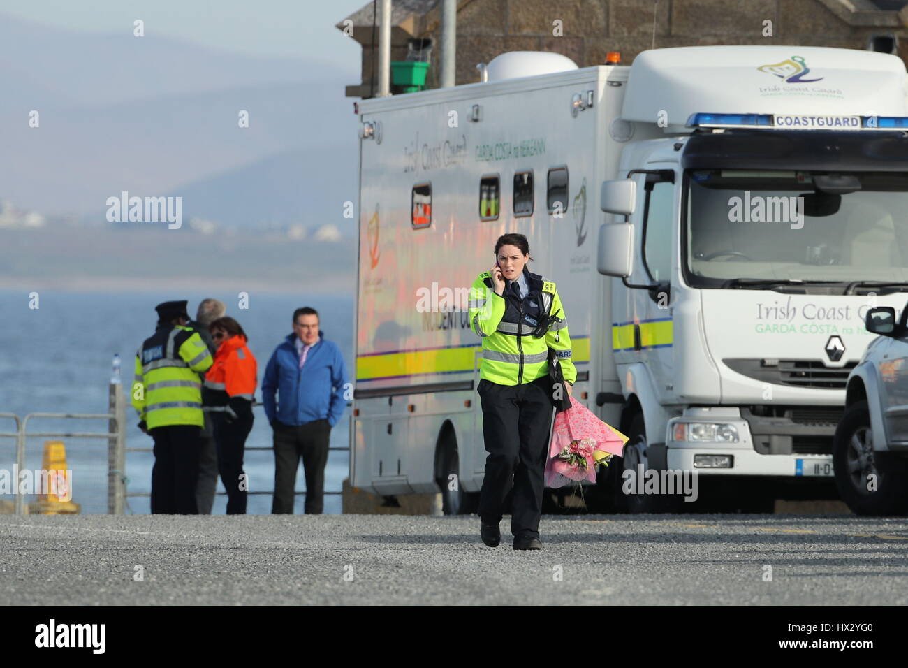 A Police Officer Walks Past An Irish Coastguard Vehicle With Flowers At