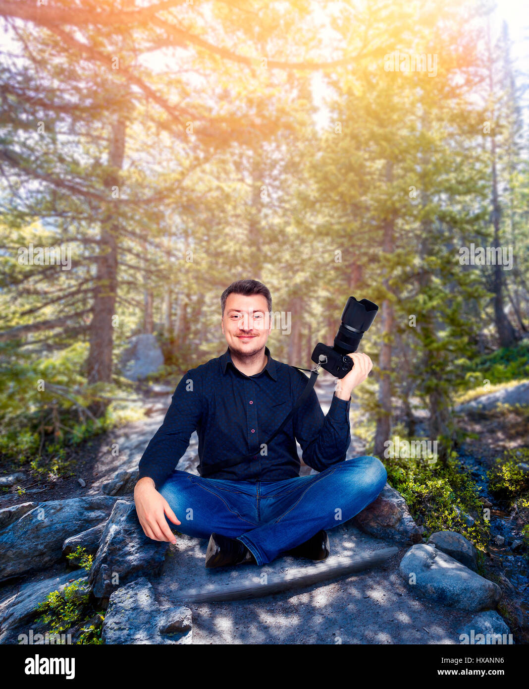 Smiling photographer with camera sitting in a yoga pose, pine forest on ...
