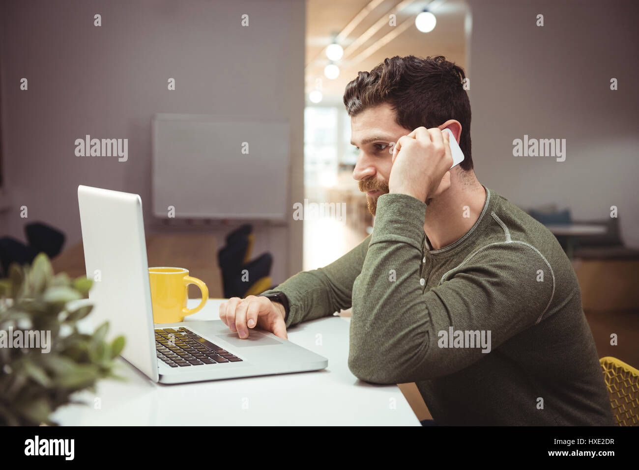 Male executive talking on mobile phone while using laptop at desk in ...