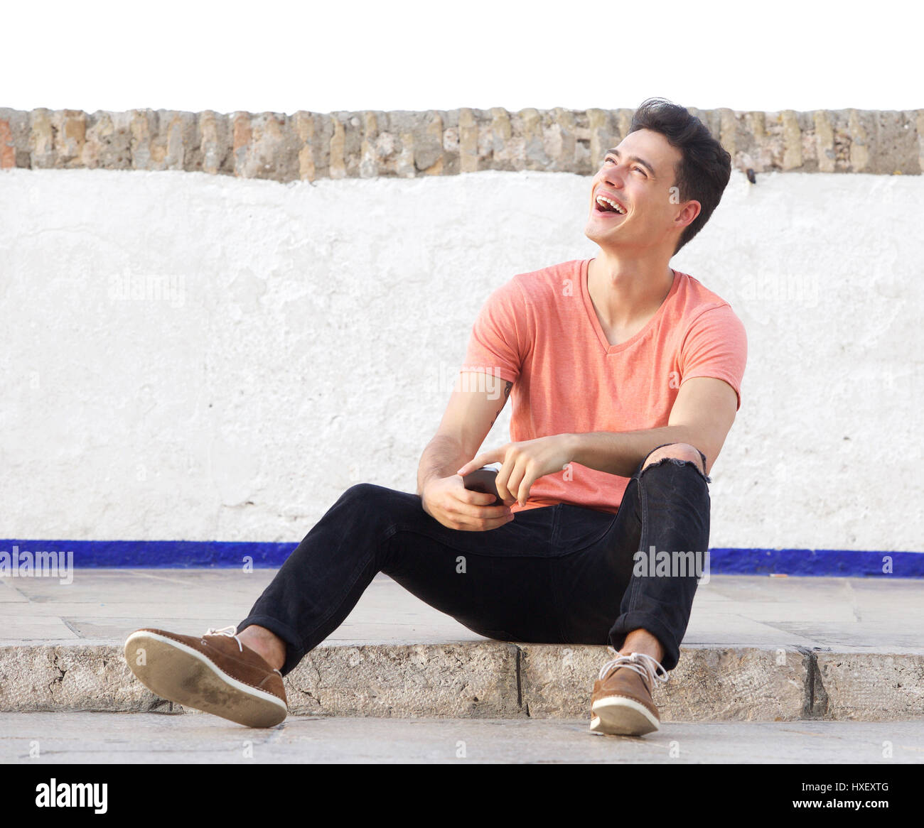 Portrait of a cheerful guy laughing and sitting outside on sidewalk ...