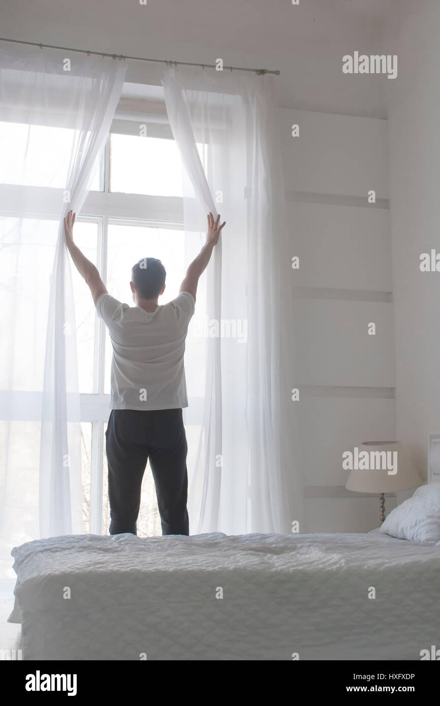 Young man opening curtains to welcomw morning and light, back view ...