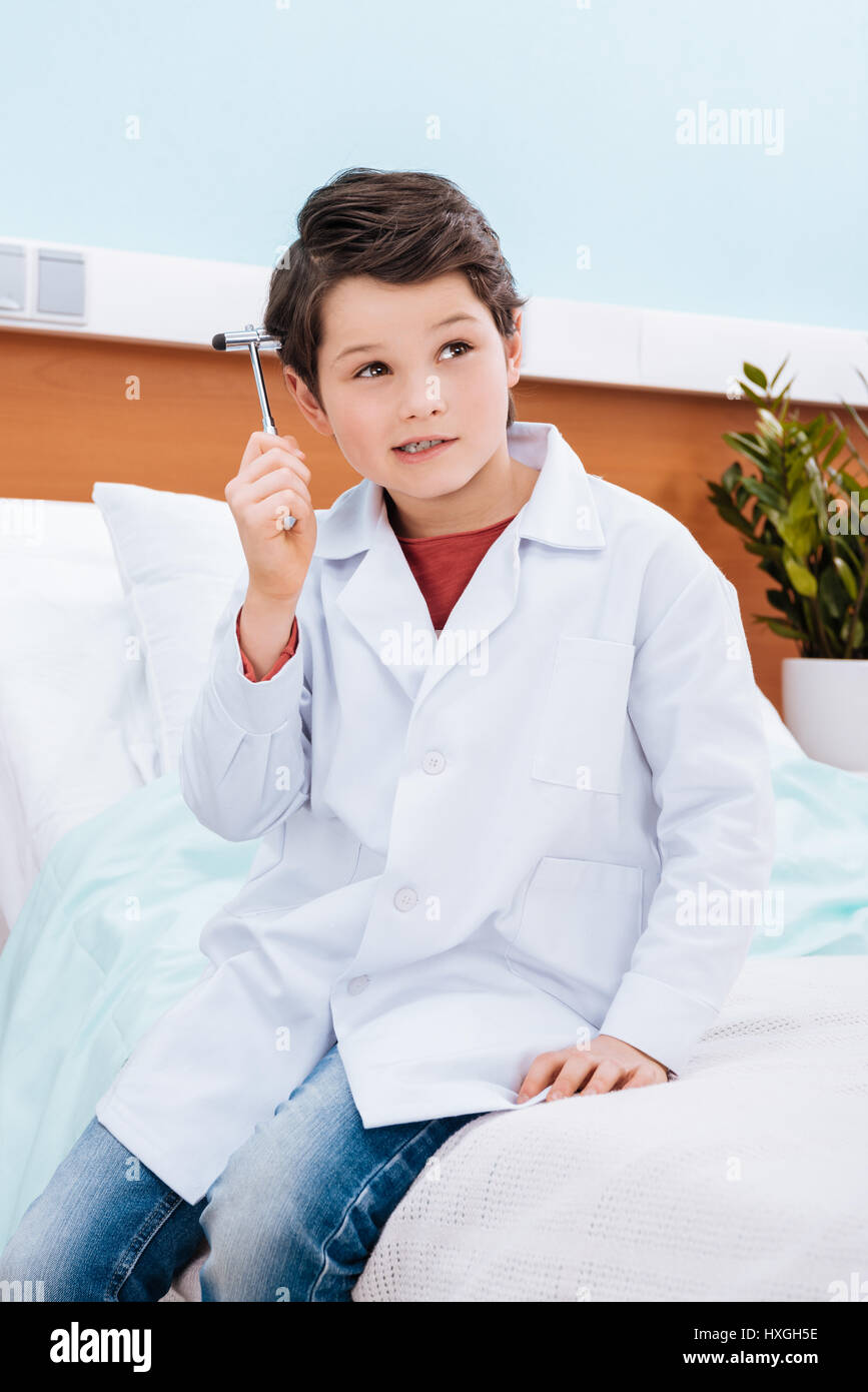 Confused boy doctor in medical uniform sitting on bed with reflex ...