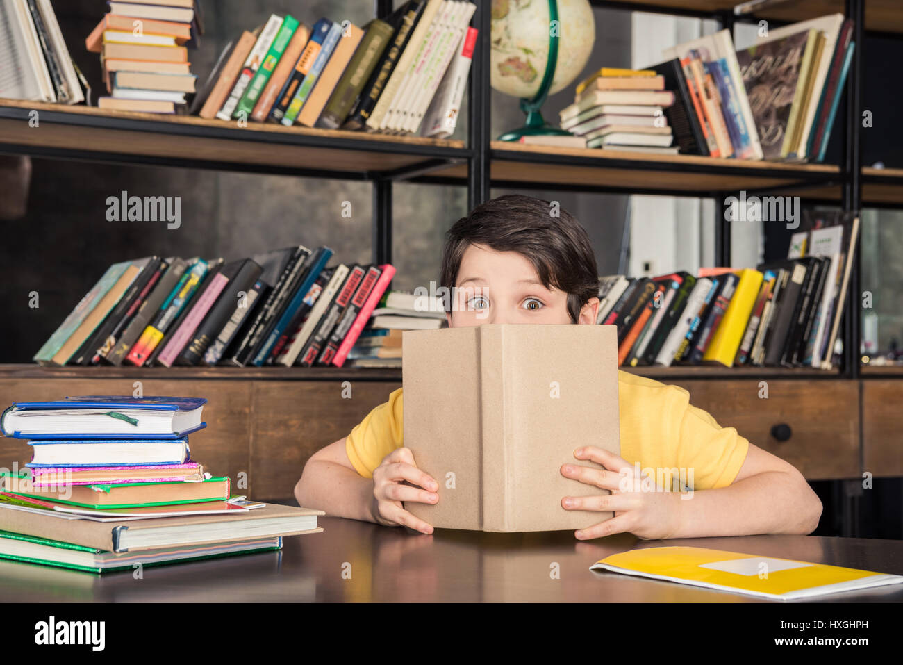Shocked schoolboy peering over book in library Stock Photo - Alamy