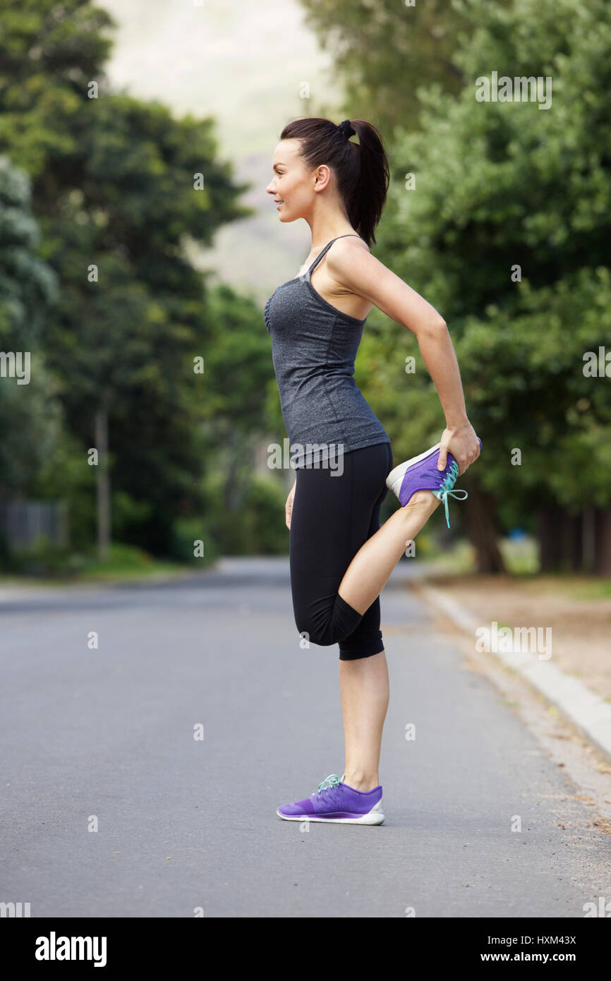 Side full body portrait of a sporty young woman doing stretching exercise outdoors in a park ...