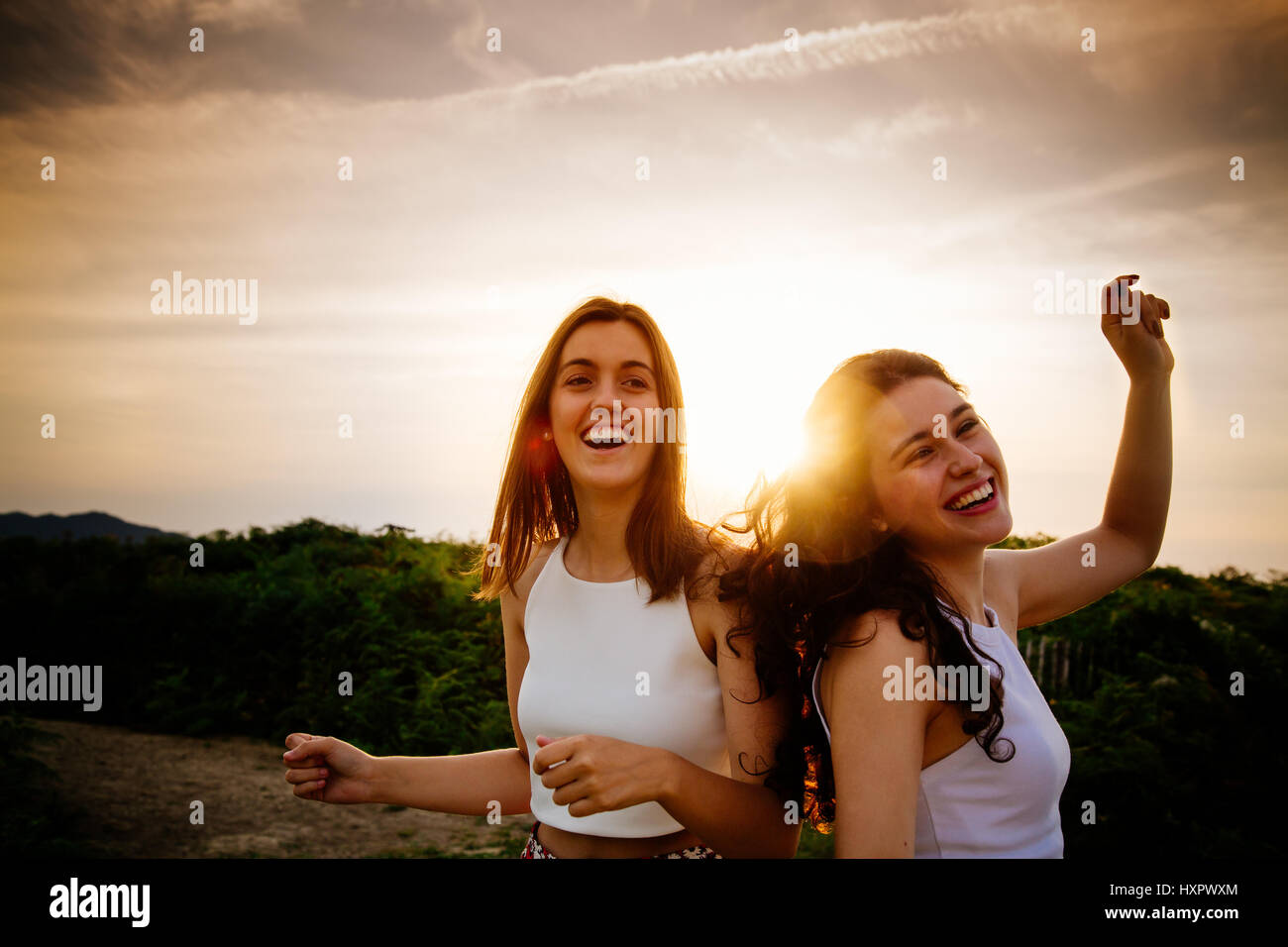 Happy and funny female friends dancing on a cliff at sunset. Sun flare ...