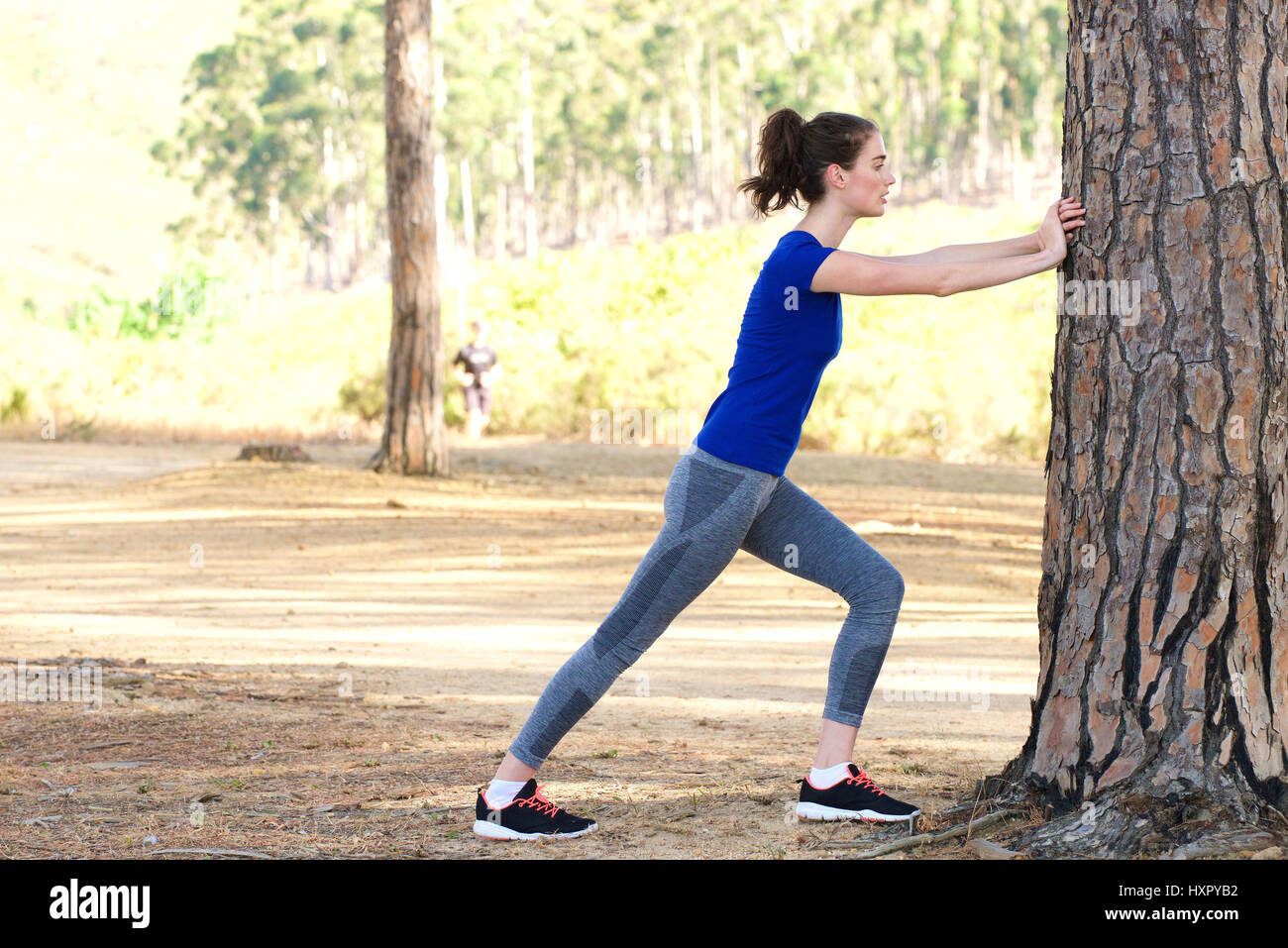 Full length side portrait of sporty woman stretching against tree ...