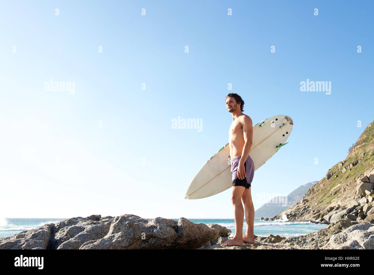 Full length portrait of attractive young man standing on beach carrying ...