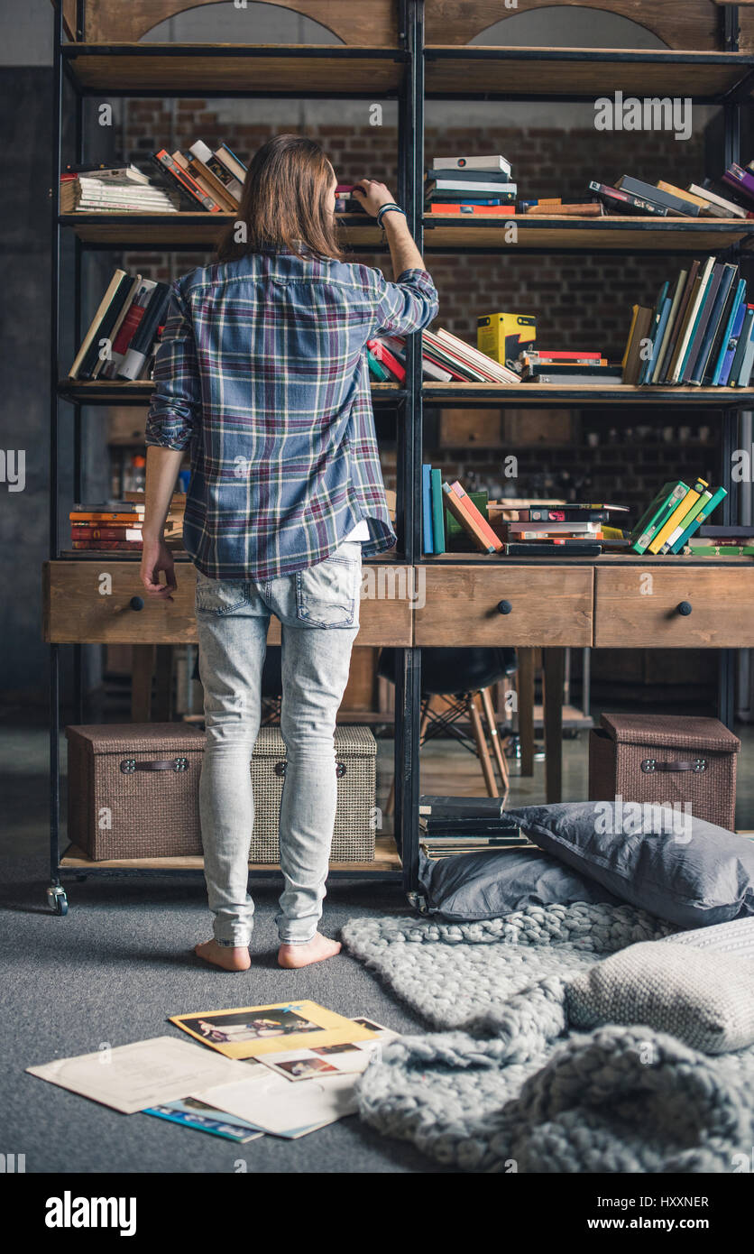 Back view of young barefoot man choosing books from bookshelves at home ...