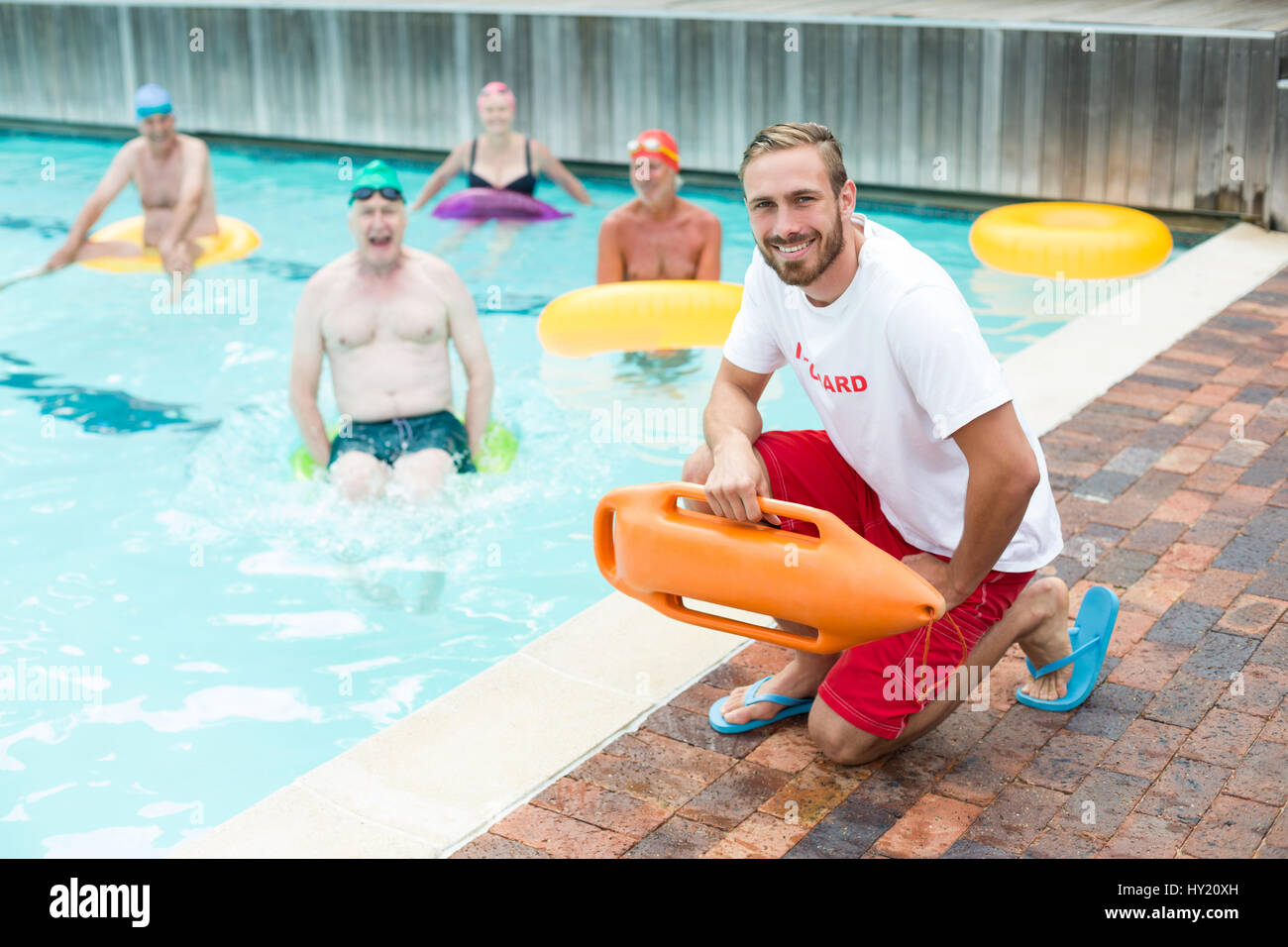 Portrait of male lifeguard crouching while swimmers swimming in pool ...